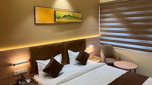 Quarto de hotel com cama, mesa de cabeceira, luminária, cadeira e janela com persianas.