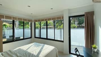 Quarto com cama, janelas amplas, porta de vidro deslizante e vista para o verde lá fora.
