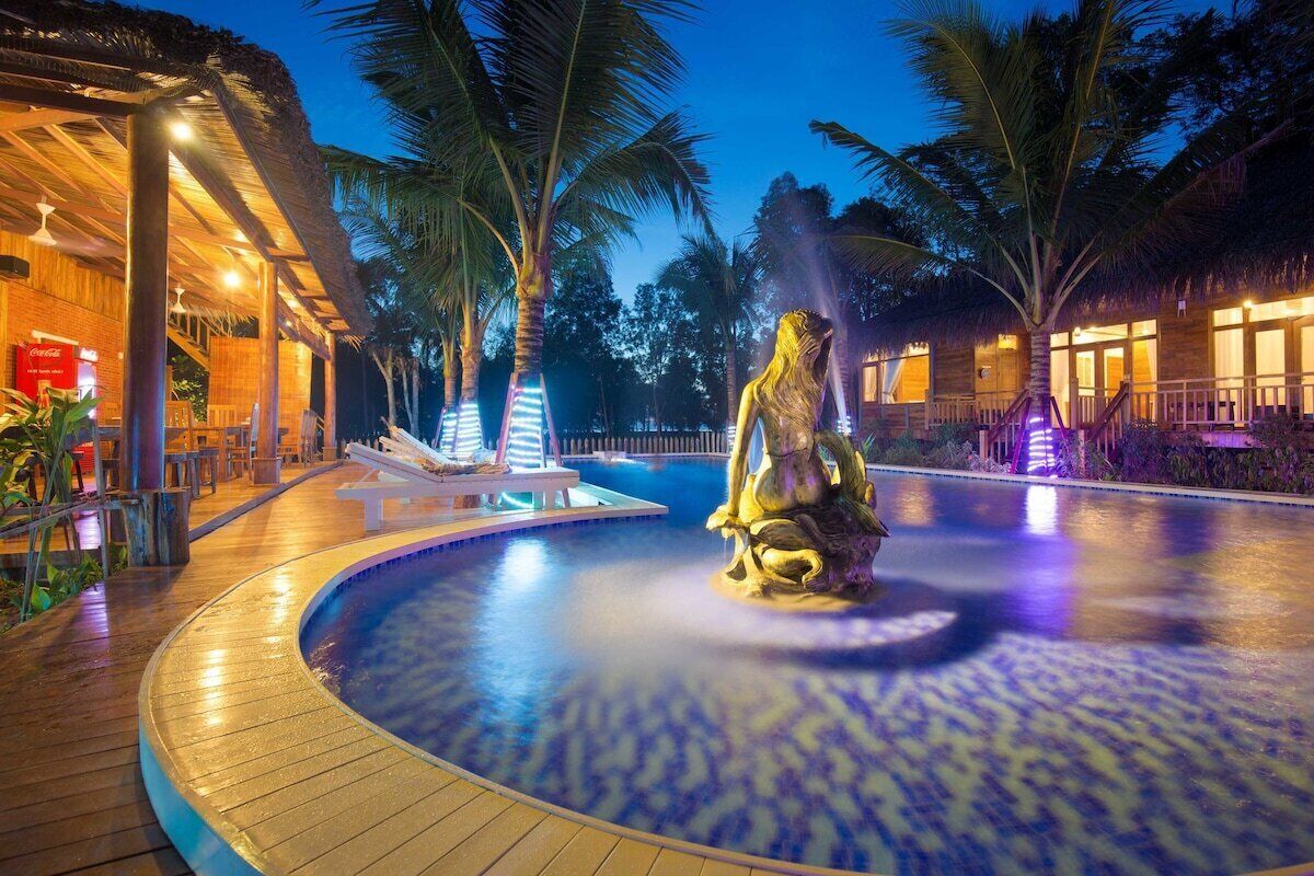 Uma área de piscina com estátua, deck de madeira e estruturas com telhado de palha.