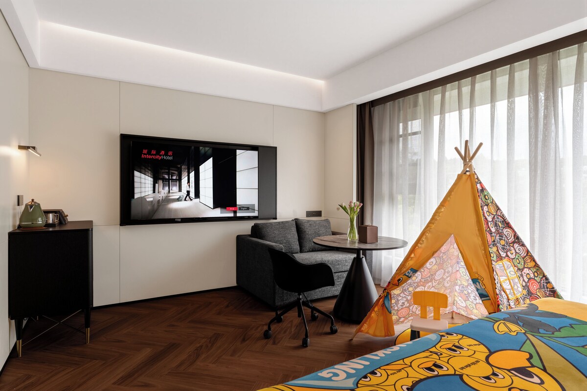 Quarto de hotel com cama, escrivaninha, cadeira, TV fixada na parede e janela com cortinas.