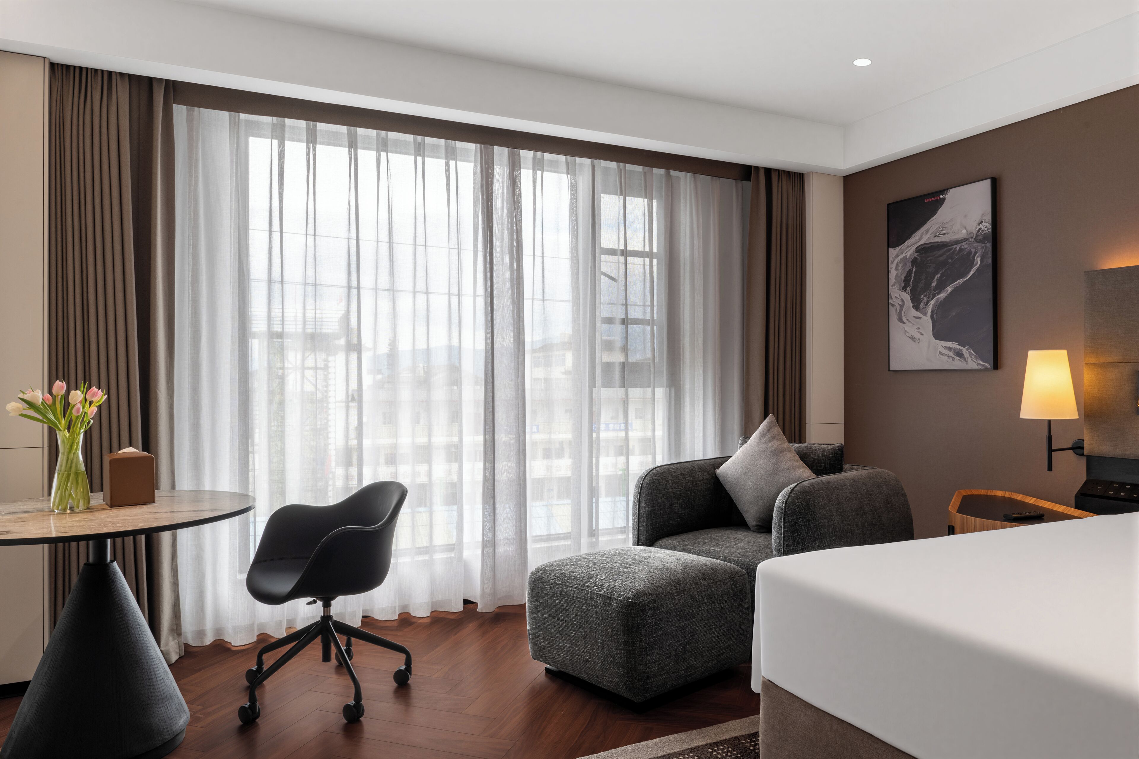 Quarto de hotel com cama, escrivaninha com cadeira, sofá, abajur e um quadro na parede.