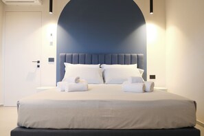 Quarto de hotel moderno com uma cama grande, cabeceira azul escura e duas mesinhas de cabeceira.