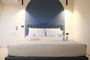 Quarto de hotel moderno com uma cama grande, cabeceira azul escura e duas mesinhas de cabeceira.