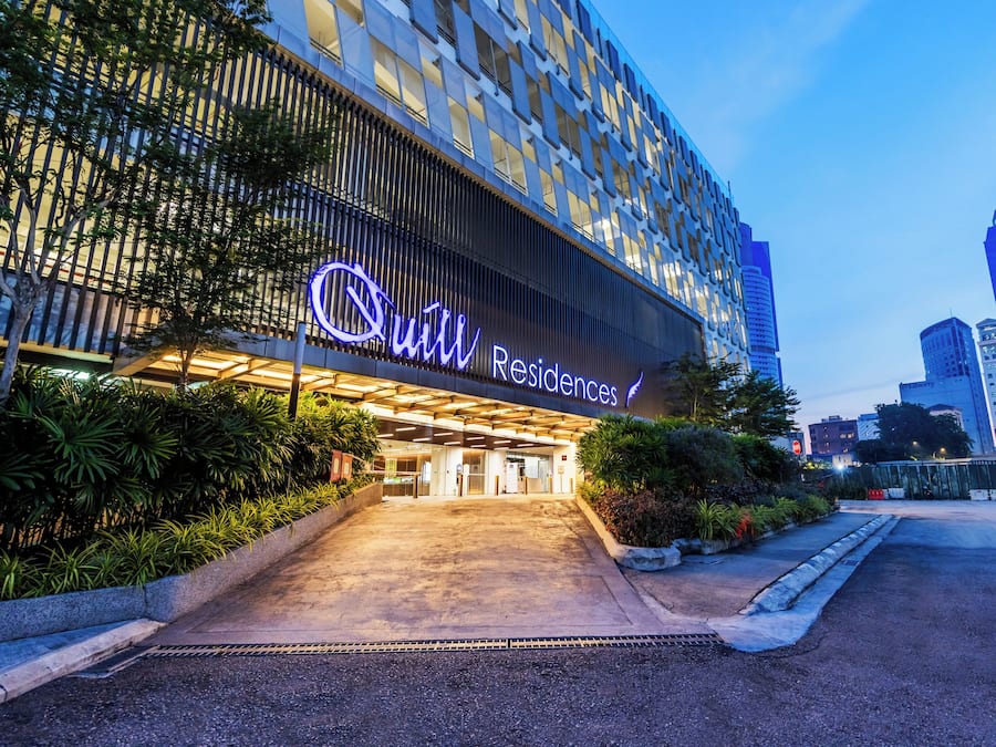 Entrada de hotel com fachada moderna, uma placa iluminada em destaque e um portão de acesso.