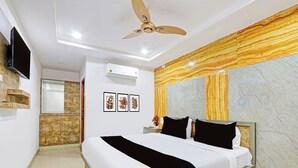 Quarto de hotel com cama, ventoinha no teto, aparelho de ar condicionado, armário de madeira, espelho e quadros nas paredes.