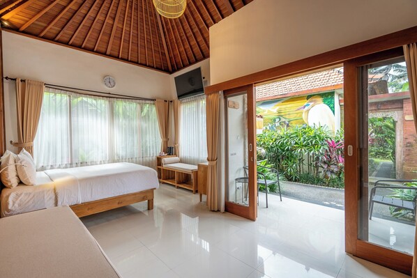 Quarto de hotel com cama, uma mesinha, uma cadeira, televisão e porta de vidro deslizante para área externa com plantas.