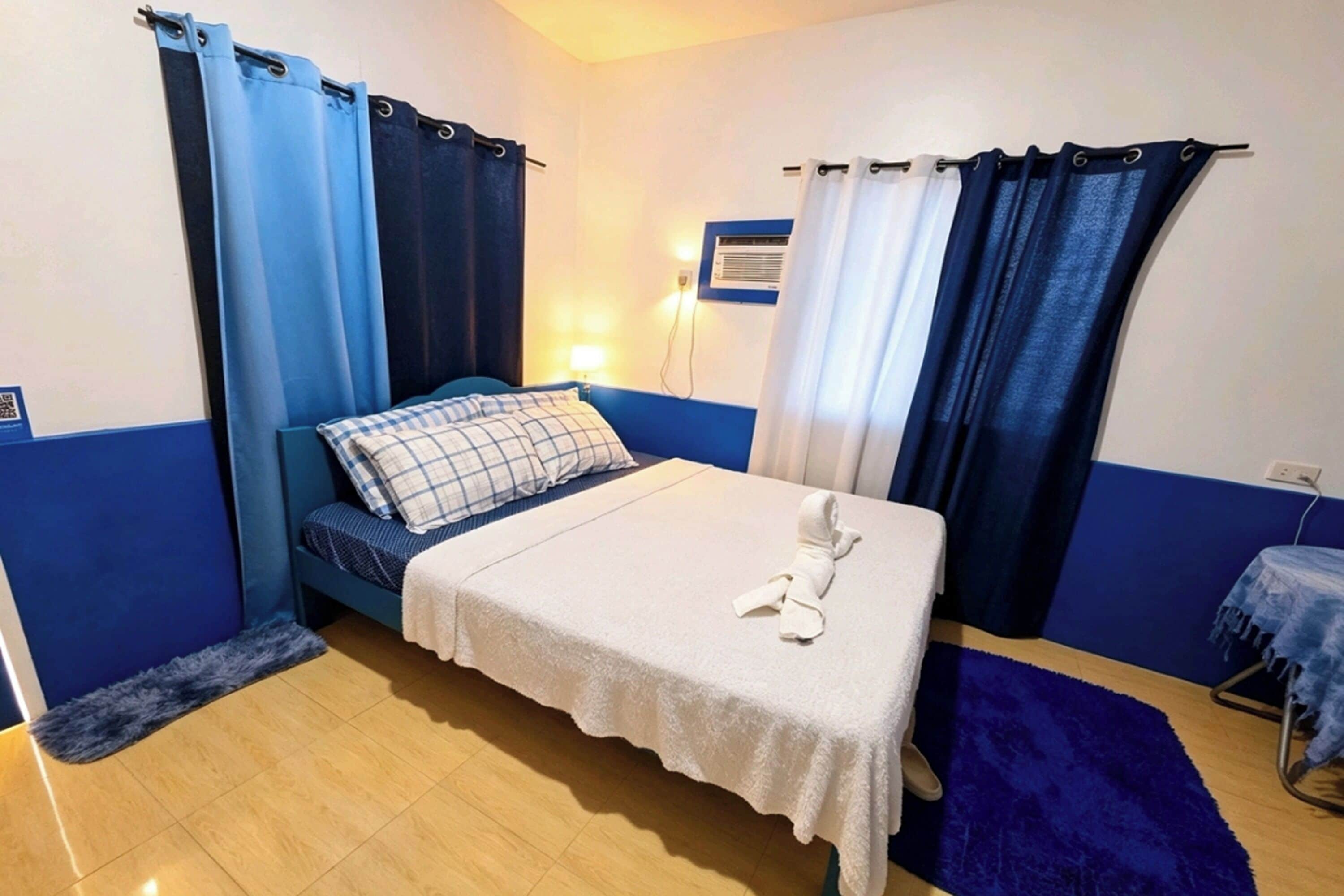 Uma cama de solteiro com colcha branca e toalha, parede azul, pequena mesa com uma cadeira e tapete azul.