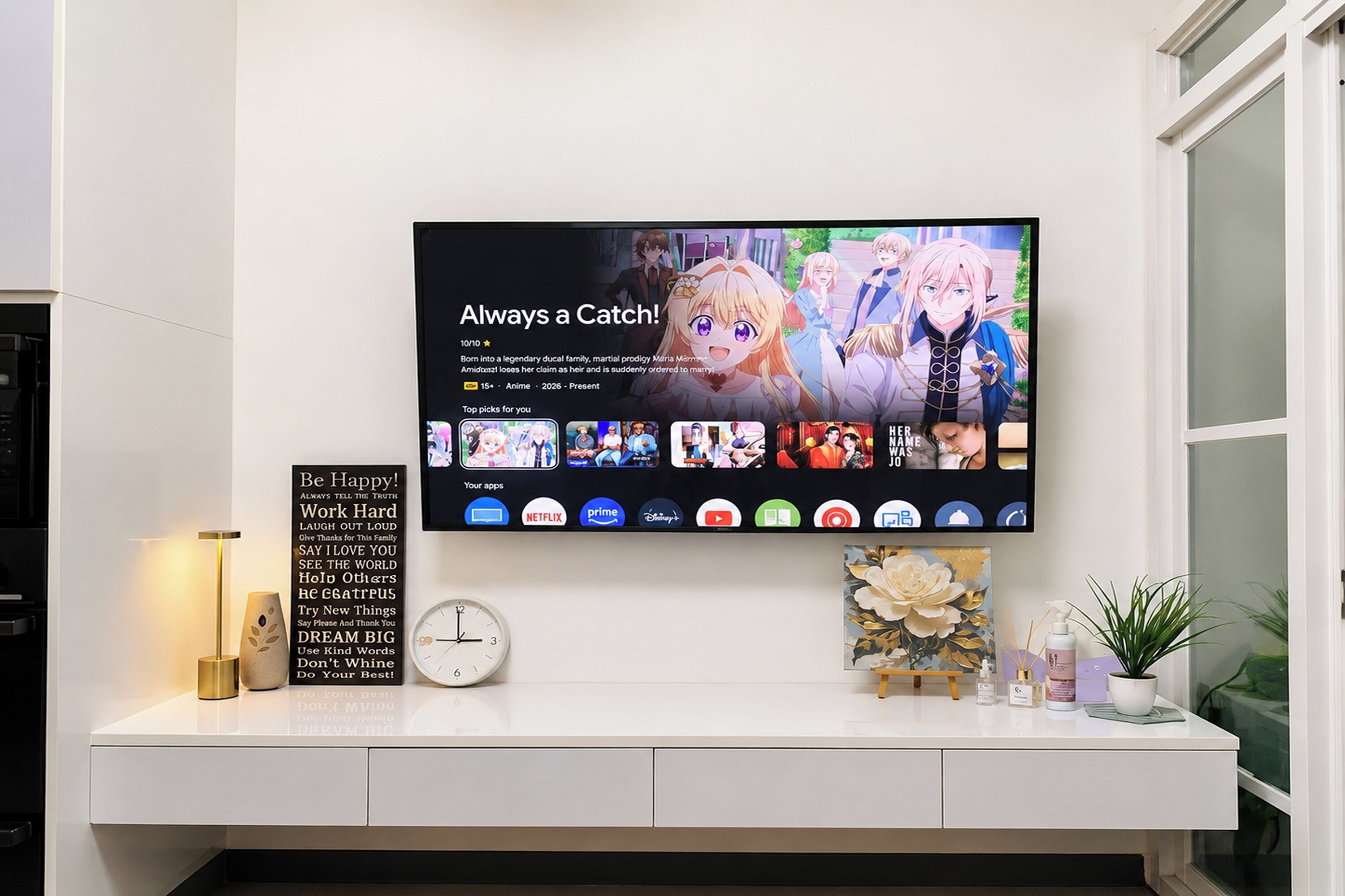 Televisão de tela plana montada em parede branca exibindo um anime com legenda. Abaixo da TV, um console branco com um relógio, uma placa com frase motivacional e um pequeno vaso de planta.