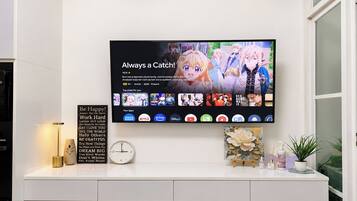 Televisão de tela plana montada em parede branca exibindo um anime com legenda. Abaixo da TV, um console branco com um relógio, uma placa com frase motivacional e um pequeno vaso de planta.