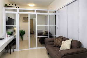 Uma sala de estar moderna com um sofá marrom, uma mesa de centro e uma TV sobre um móvel. Há uma porta de correr que dá para uma área de cozinha com uma mesa de jantar e cadeiras.