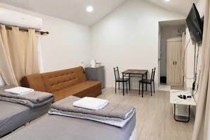 Quarto compacto com cama, sofá, pequena mesa com cadeiras e televisão na parede.