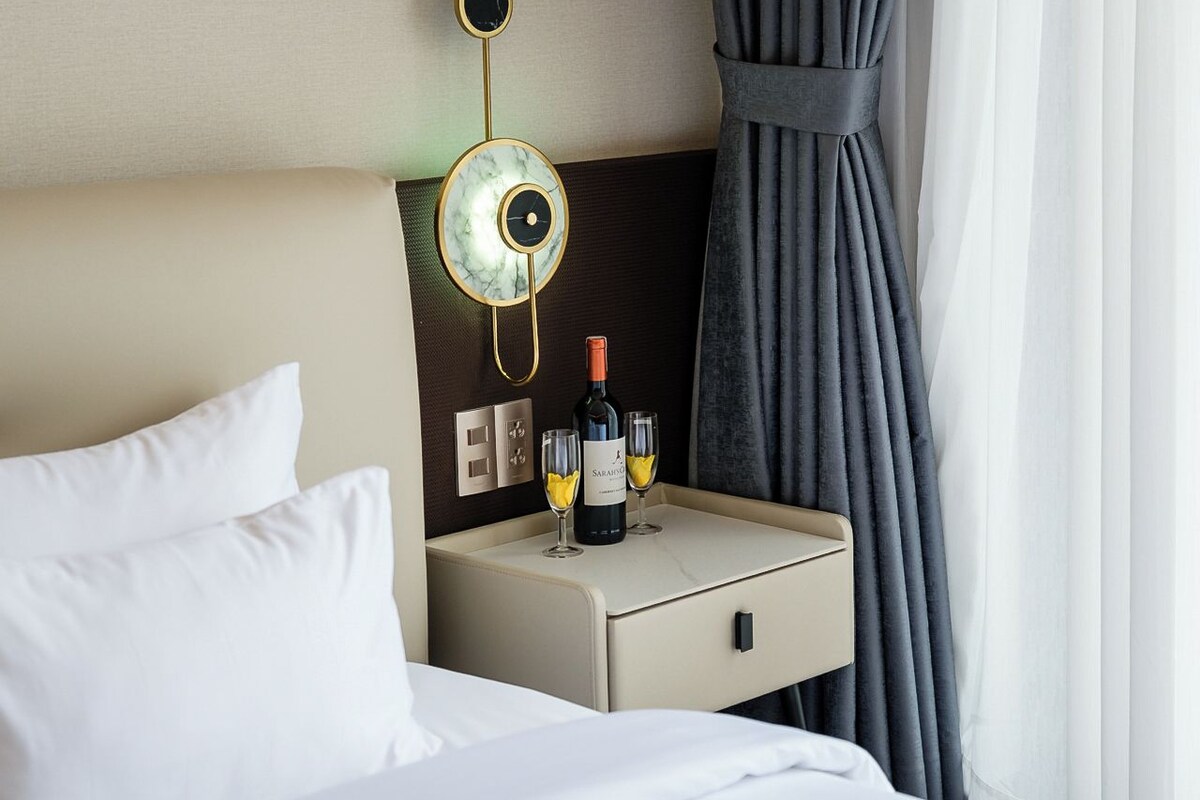 Quarto de hotel com cama, mesa de cabeceira com uma garrafa de vinho e dois copos, relógio na parede e cortina.