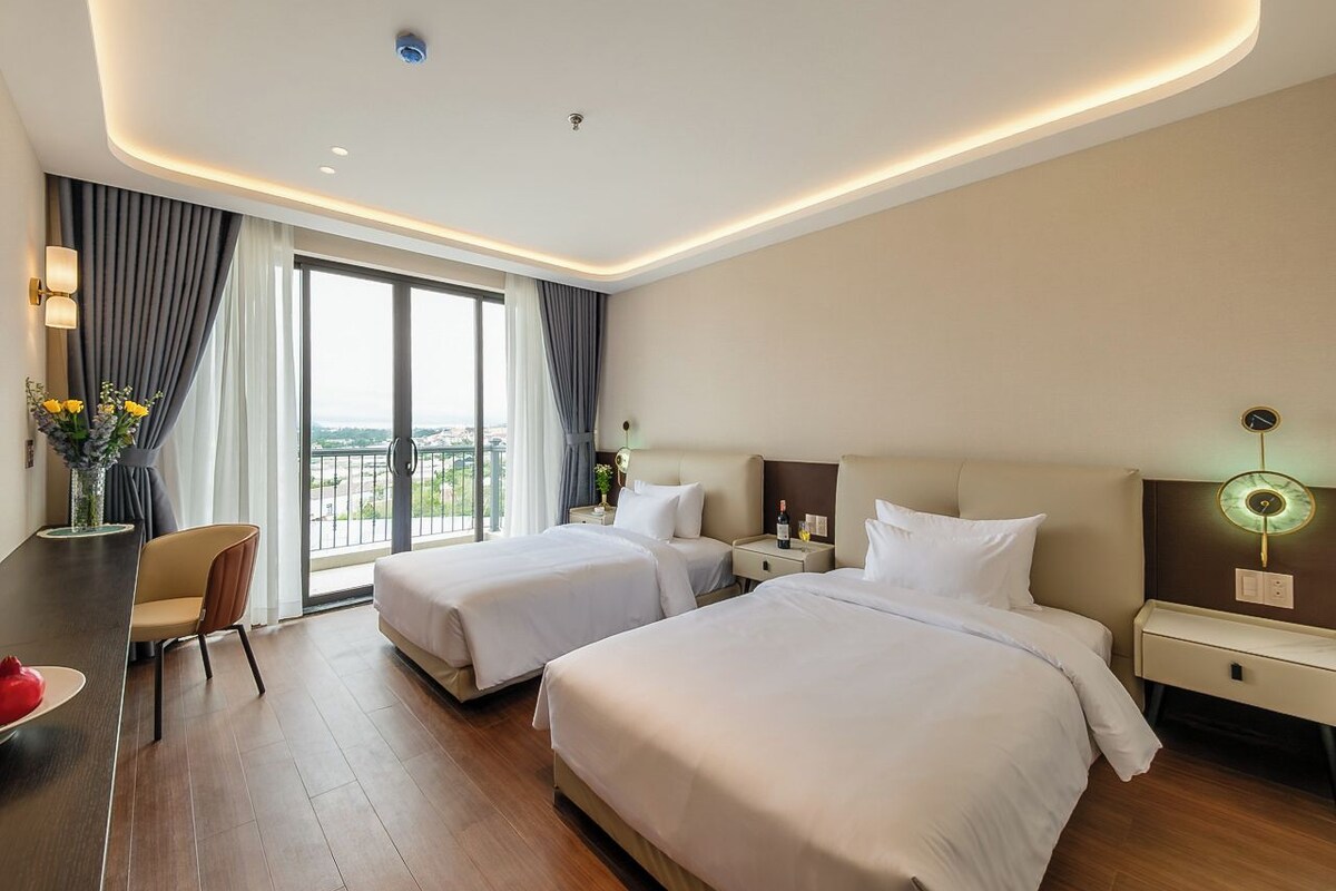 Quarto de hotel com duas camas, uma escrivaninha e vista através de uma grande janela.