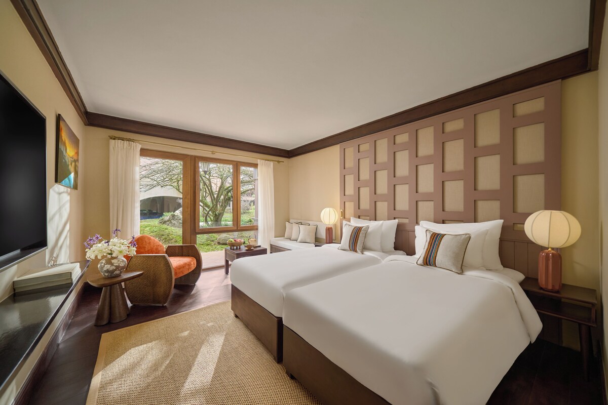 Quarto de hotel com uma cama grande, um sofá, uma TV e uma janela com vista para vegetação.