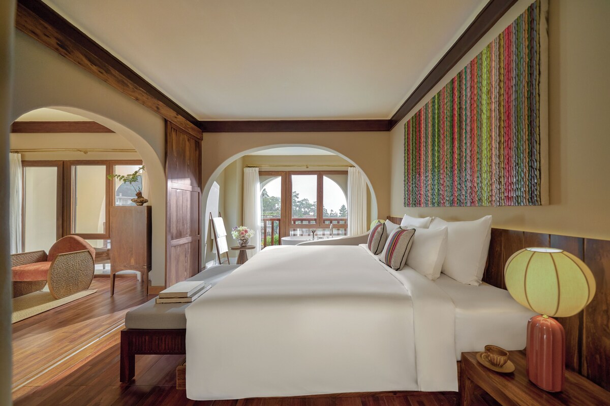 Quarto de hotel com uma cama grande, piso de madeira, vista através de uma porta arqueada e uma obra de arte colorida na parede.