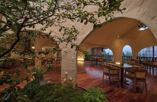 Um interior espaçoso com tetos altos em arco, piso de madeira e uma área de estar central com mesas e cadeiras.