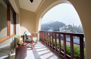Uma varanda com uma cadeira de madeira, uma mesinha com uma planta e vista para uma colina com edifícios.