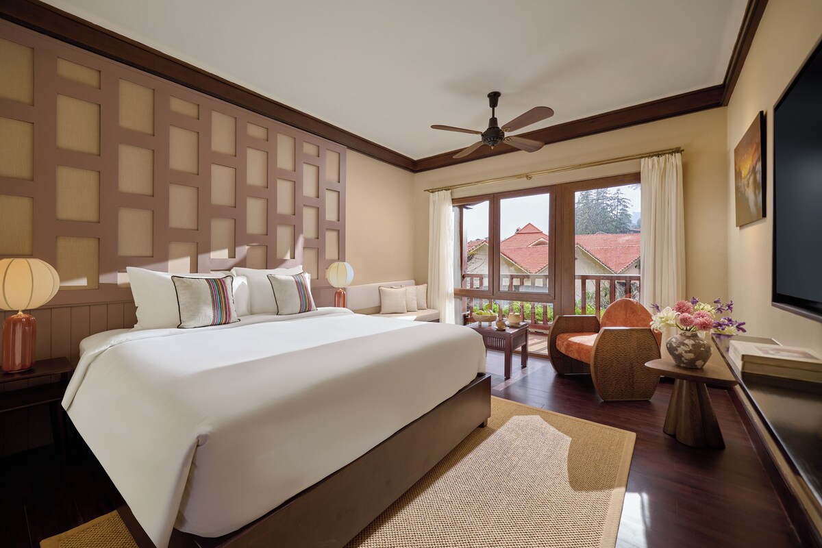 Quarto de hotel com uma cama grande, ventilador de teto, área de estar com mesa e cadeiras, e vista para prédios pela janela.