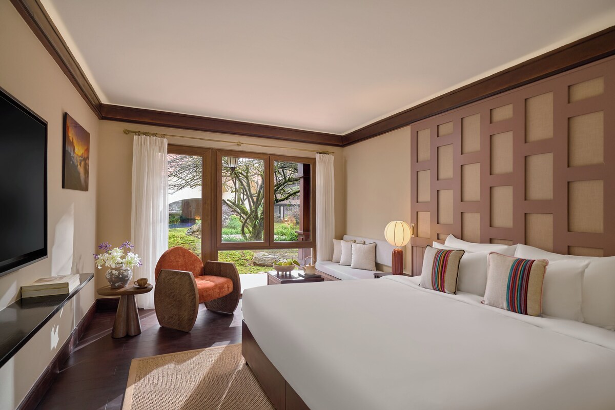 Quarto de hotel com uma cama grande, televisão, área de estar com uma poltrona e vista para o exterior.