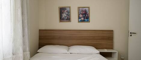 Quarto de hotel com cabeceira de madeira, cama com roupa de cama branca, dois quadros florais emoldurados na parede e uma porta branca.