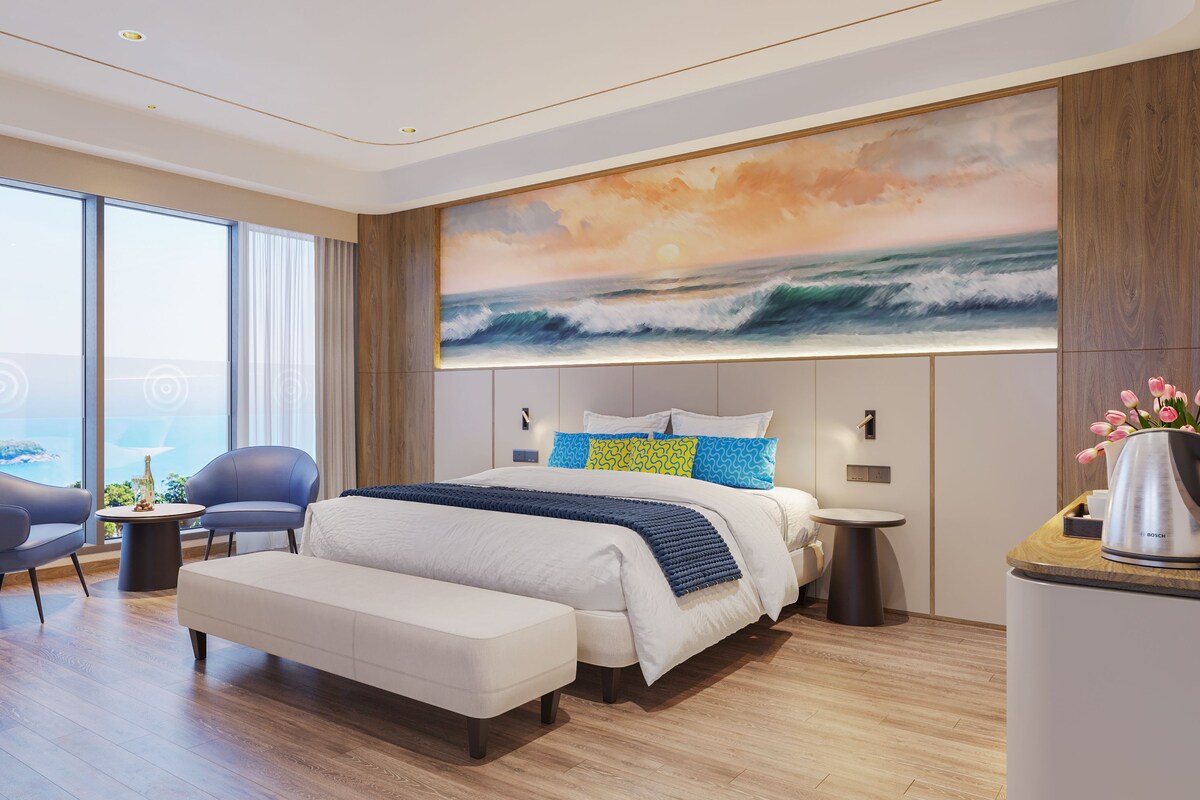 Quarto de hotel moderno com uma cama grande, área de estar com duas cadeiras, vista para o oceano através de janelas amplas e uma pintura de ondas na parede.
