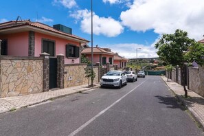 Uma rua residencial com casas, um muro de pedra e carros estacionados.
