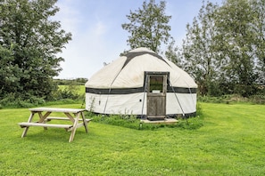 Uma yurta com porta, uma mesa de piquenique e um campo gramado com árvores ao fundo.