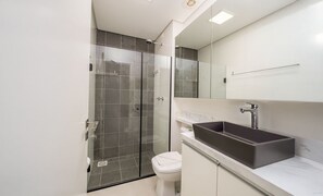 Uma casa de banho moderna com cabine de duche em vidro, sanita branca e uma grande pia retangular com bancada de mármore.