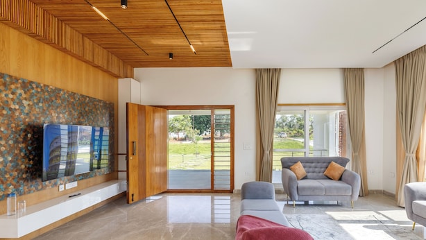 Uma sala de estar moderna com teto de madeira, uma televisão grande, um sofá cinza e vista para um jardim.