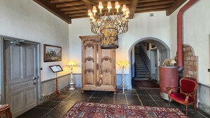 Um corredor imponente com um candelabro, uma cadeira vermelha e uma escadaria que leva a outro cômodo.