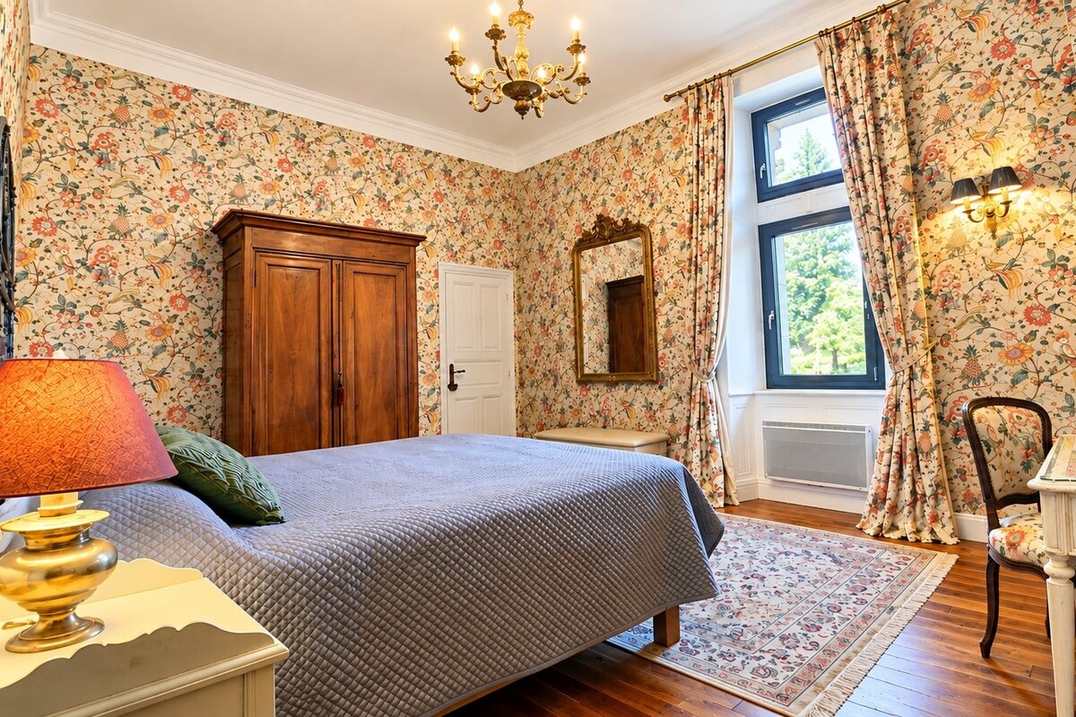 Quarto com cama, mesa de cabeceira, cômoda, espelho, janela com cortinas e lustre.