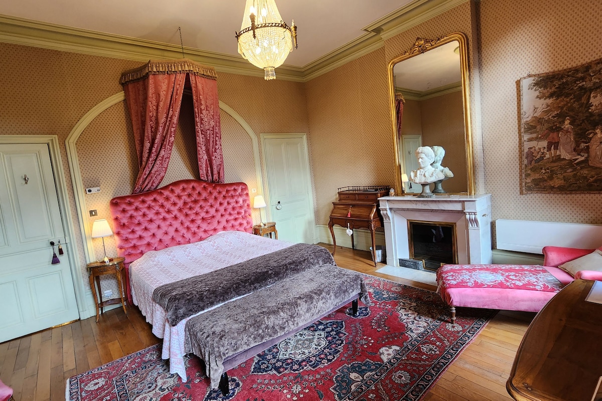 Quarto com cama grande, lareira, espelho e cadeira vermelha.