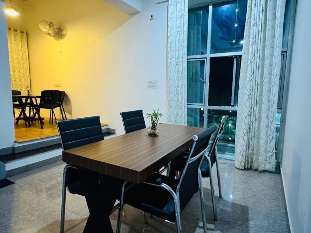 Uma sala de jantar com uma mesa e cadeiras de madeira, uma planta em vaso e vista pela janela com cortinas.