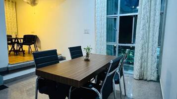 Uma sala de jantar com uma mesa e cadeiras de madeira, uma planta em vaso e vista pela janela com cortinas.