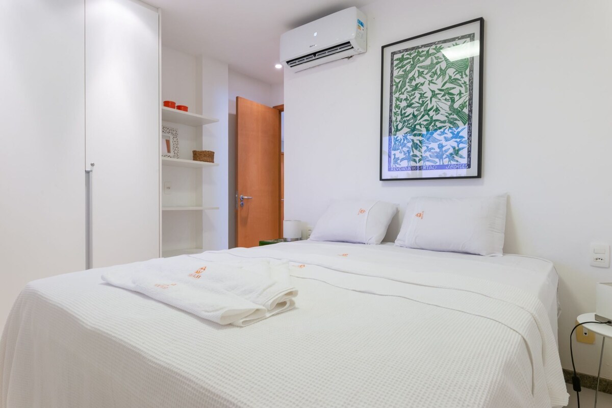 Quarto de hotel com uma cama, um guarda-roupa, uma prateleira com recipientes vermelhos, um aparelho de ar condicionado e uma obra de arte emoldurada na parede.