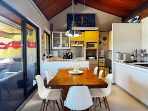 Cozinha moderna com mesa de jantar de madeira e cadeiras brancas, luminária pendente e vista para o exterior através de uma porta de vidro.