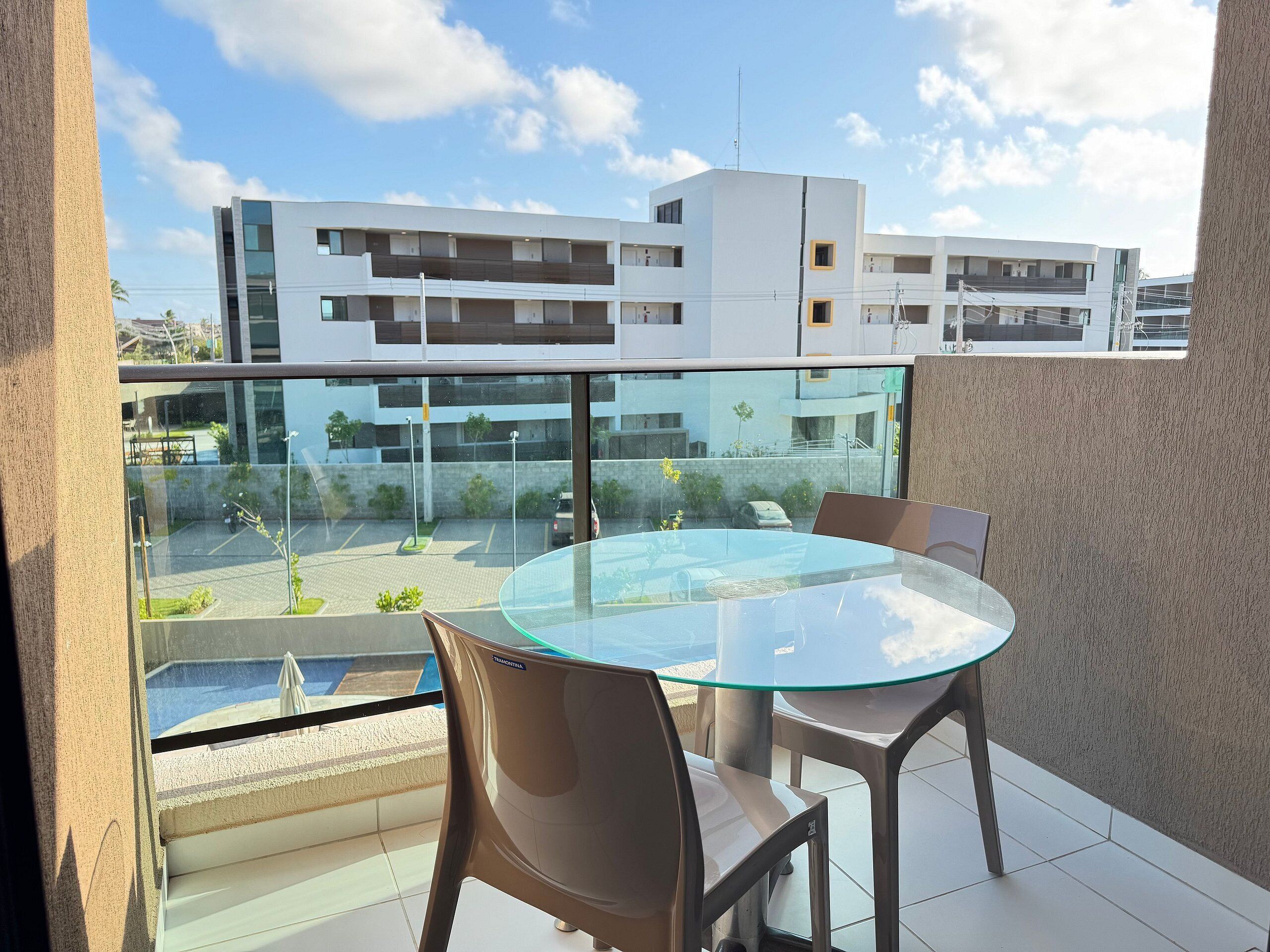 Uma varanda com mesa e cadeiras de vidro, com vista para um estacionamento e edifícios modernos.
