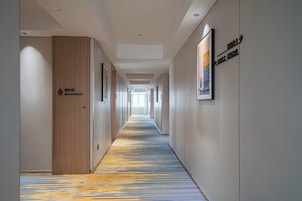 Corredor de hotel com carpete estampado, portas de madeira e quadros nas paredes.