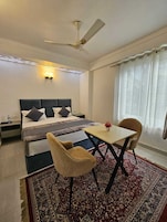 Um quarto com cama, mesa de cabeceira, tapete, um ventilador no teto, uma mesa com duas cadeiras e uma janela com cortinas.