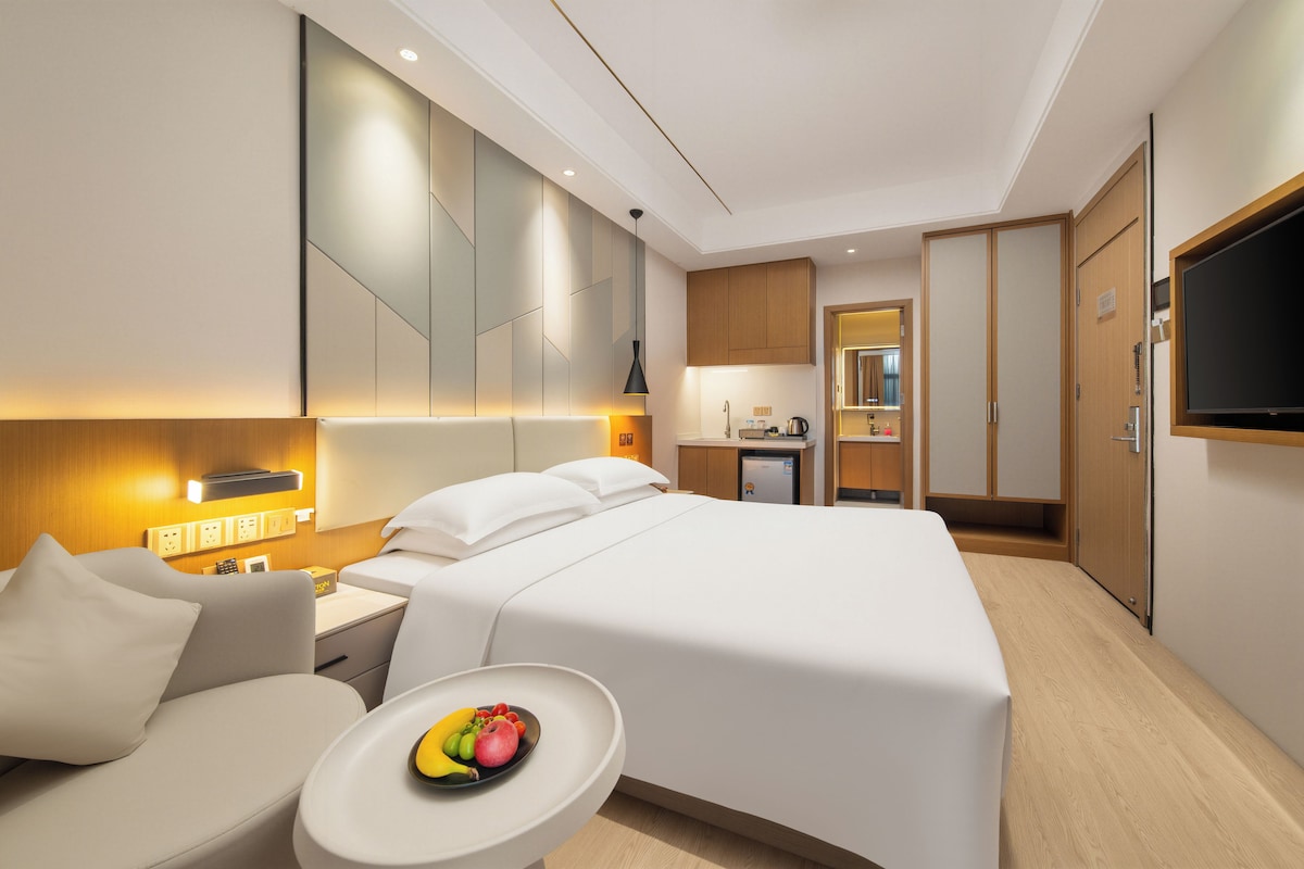 Quarto de hotel com cama grande, mesa de cabeceira com cesta de frutas, sofá, pequeno espaço de cozinha e televisão.
