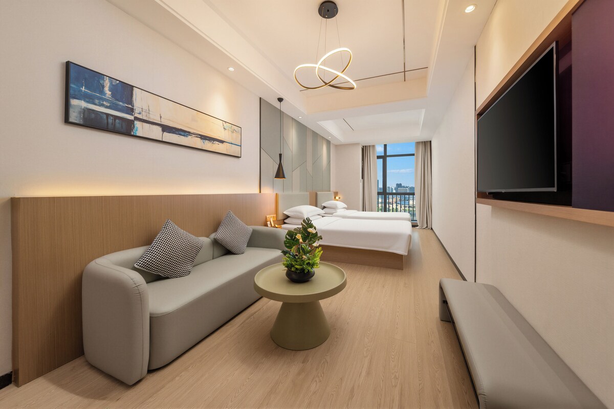 Quarto de hotel moderno com sofá, uma pequena mesa com uma planta, cama, televisão e vista para a cidade.