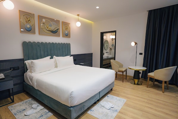 Quarto de hotel com cama grande, mesa de cabeceira, uma cadeira, uma mesinha e uma obra de arte na parede.