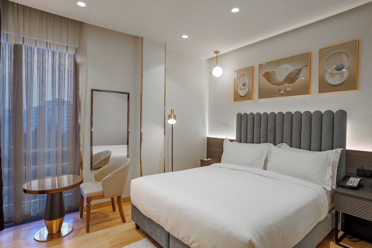 Quarto de hotel moderno com cama, mesa de cabeceira, cadeira e uma janela grande com cortinas.