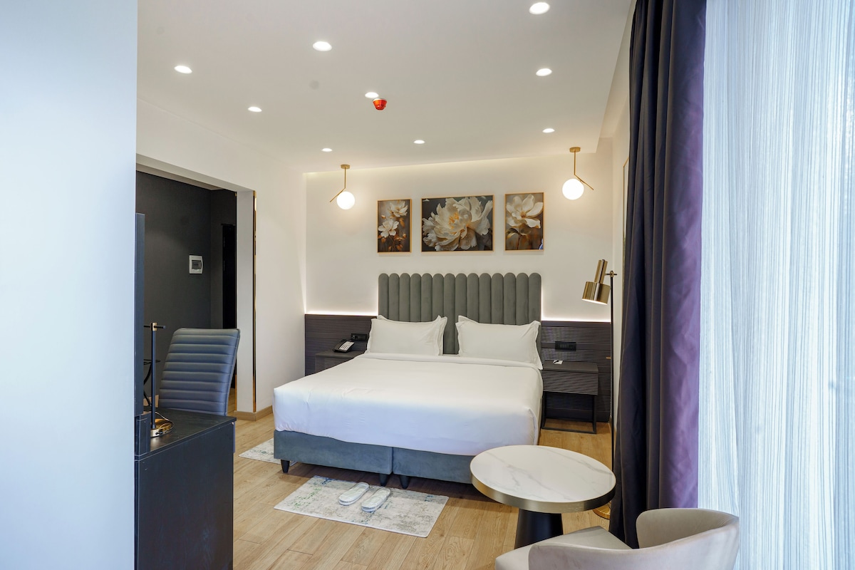 Quarto de hotel moderno com uma cama grande, uma escrivaninha, uma cadeira e uma mesa redonda.