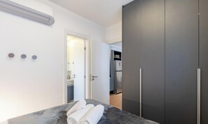 Quarto moderno com uma cama grande, um guarda-roupa e uma kitchenette ao fundo.