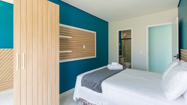 Quarto moderno com cama, guarda-roupa de madeira e banheiro visível por uma porta aberta.