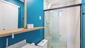 Um banheiro moderno com parede azul, box de vidro, vaso sanitário branco e prateleira de madeira com artigos de higiene pessoal.