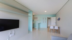 Uma sala de estar moderna com uma TV de tela plana na parede, uma área de jantar com mesa e cadeiras, e uma cozinha ao fundo.