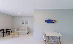 Uma sala de estar moderna com um sofá branco, cadeiras pretas, uma mesa de centro de madeira e uma mesa de jantar branca com cadeiras. Há duas imagens emolduradas na parede.