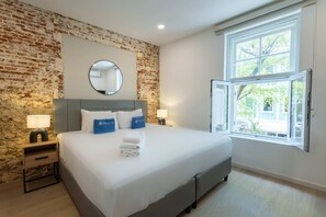 Quarto de hotel com parede de tijolos, uma cama grande com cabeceira cinza, duas mesas de cabeceira com luminárias, um espelho redondo e uma janela com vista aberta.